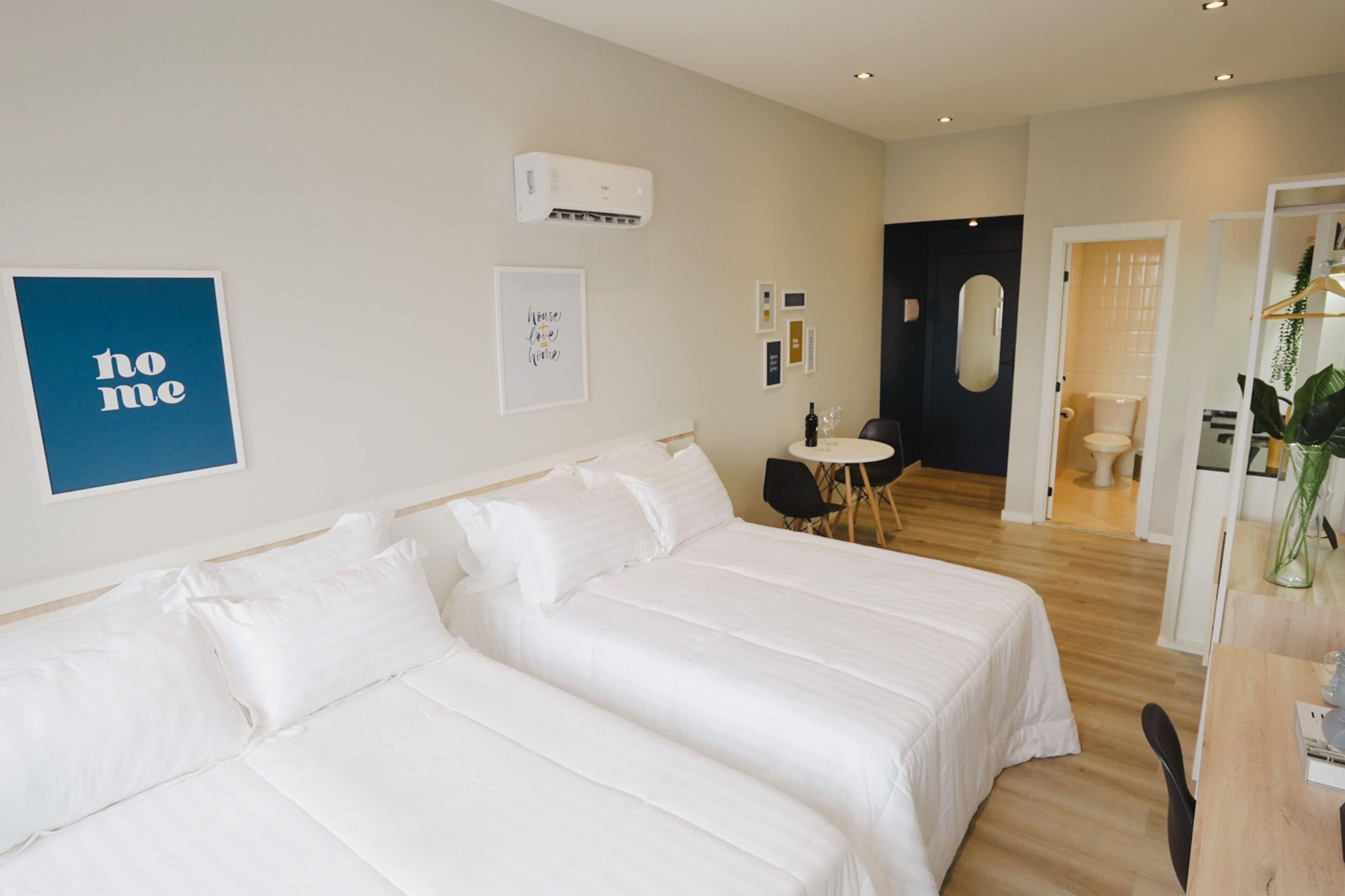 Quarto de hotel com duas camas, uma mesinha, uma cadeira e um espelho.