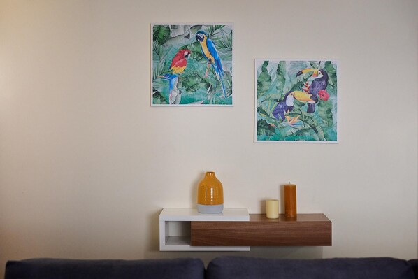 Quarto com parede branca, prateleira branca com um vaso e velas, e duas pinturas de pássaros coloridos.