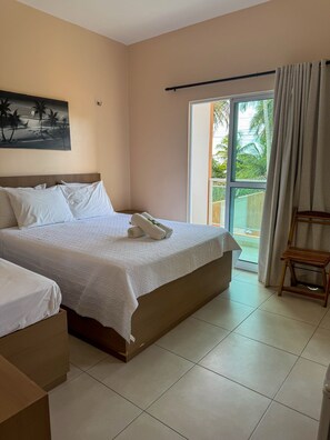 Quarto de hotel com uma cama grande, uma mesa pequena, uma cadeira e uma varanda com cortinas.