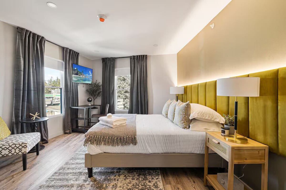 Quarto de hotel moderno com uma cama grande, uma escrivaninha, uma cadeira, uma televisão e vista para o exterior.