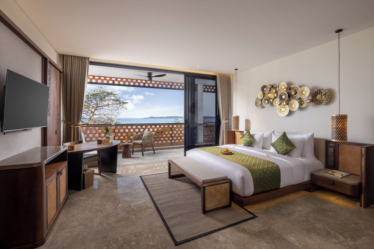 Quarto de hotel moderno com uma cama grande, uma escrivaninha e uma televisão. Possui uma varanda com vista para o mar.