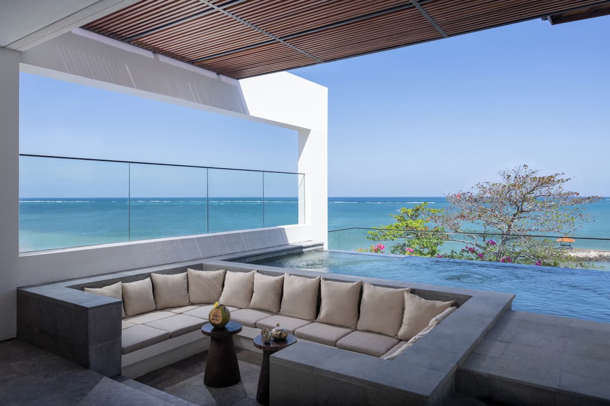 Uma piscina infinita moderna com vista clara para o oceano, um sofá bege e uma mesinha com garrafa e copos.