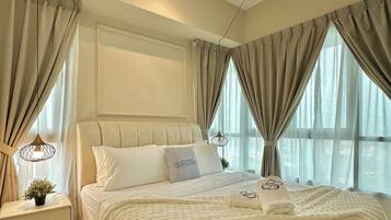 Uma cama bem arrumada com edredom branco e travesseiros, uma mesa de cabeceira com abajur e uma janela com cortinas.