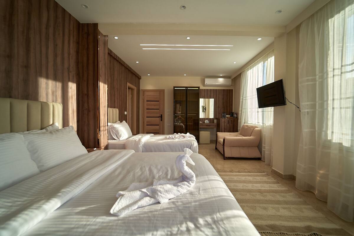 Quarto de hotel com duas camas, área de estar, televisão e uma janela grande.