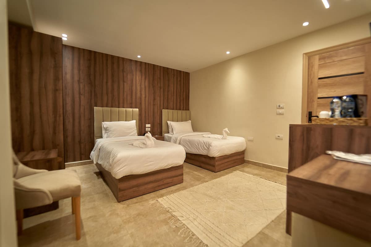 Quarto de hotel moderno com duas camas, cabeceira de madeira, uma pequena escrivaninha e uma cadeira.