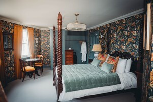 Uma cama com dossel com um edredom verde, uma cadeira de madeira, uma mesa redonda pequena e um penteado com espelho.