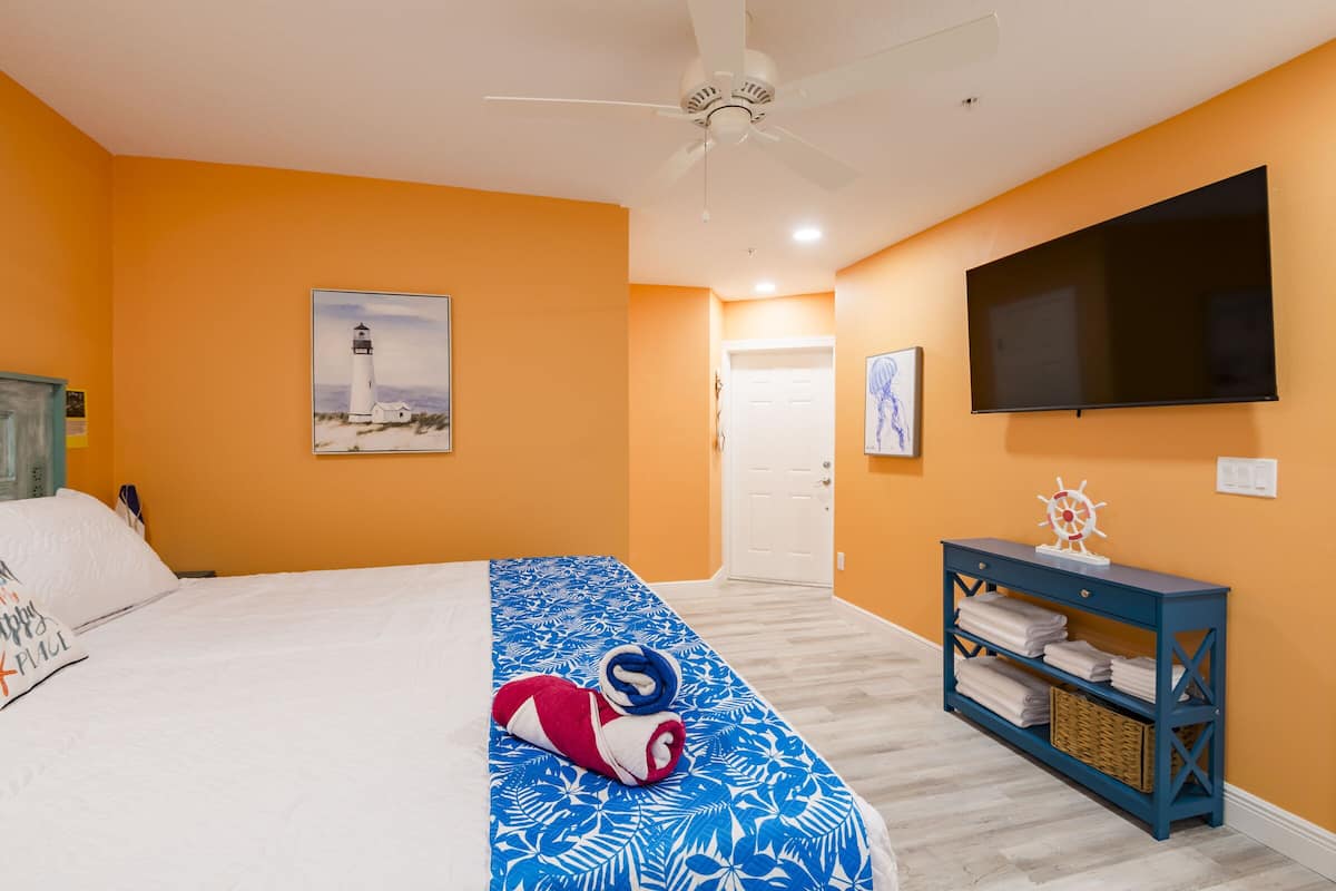 Quarto de hotel com cama, ventilador de teto, TV na parede e mesa de apoio com toalhas.