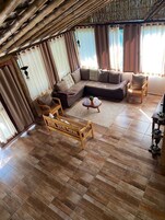 Uma sala de estar espaçosa com um grande sofá em formato de L, uma mesa de centro de madeira e um banco de madeira. O ambiente possui piso de azulejos e teto alto com vigas aparentes.
