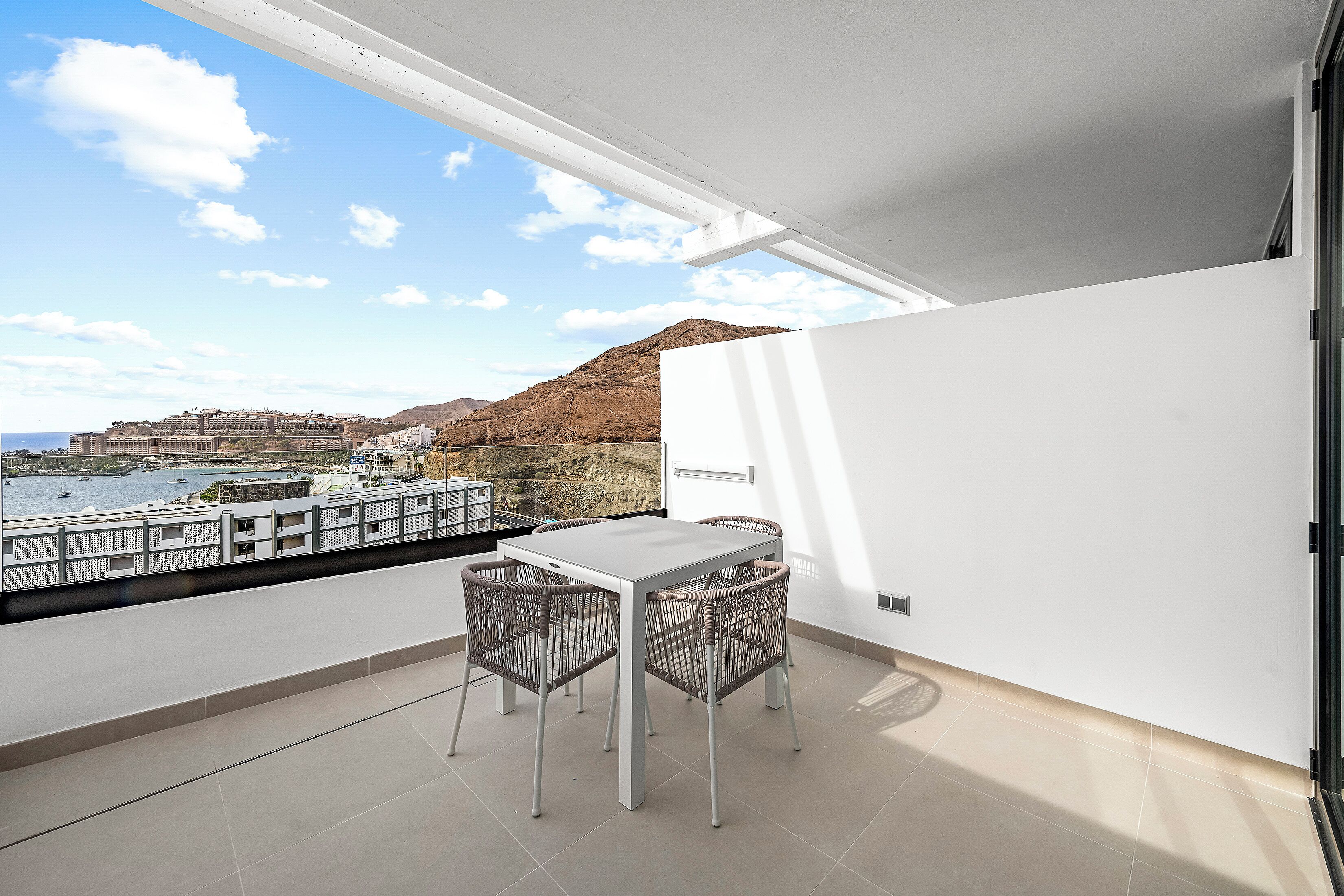 Uma varanda com uma mesa branca e cadeiras de vime, com vista para uma paisagem urbana costeira com edifícios e uma colina.