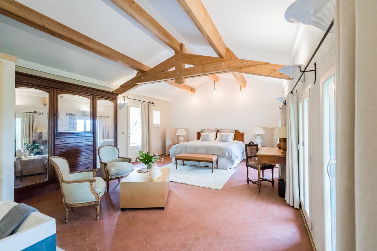 Um quarto espaçoso com uma cama grande, vigas de madeira, uma área de estar com cadeiras, uma escrivaninha e um espelho grande.
