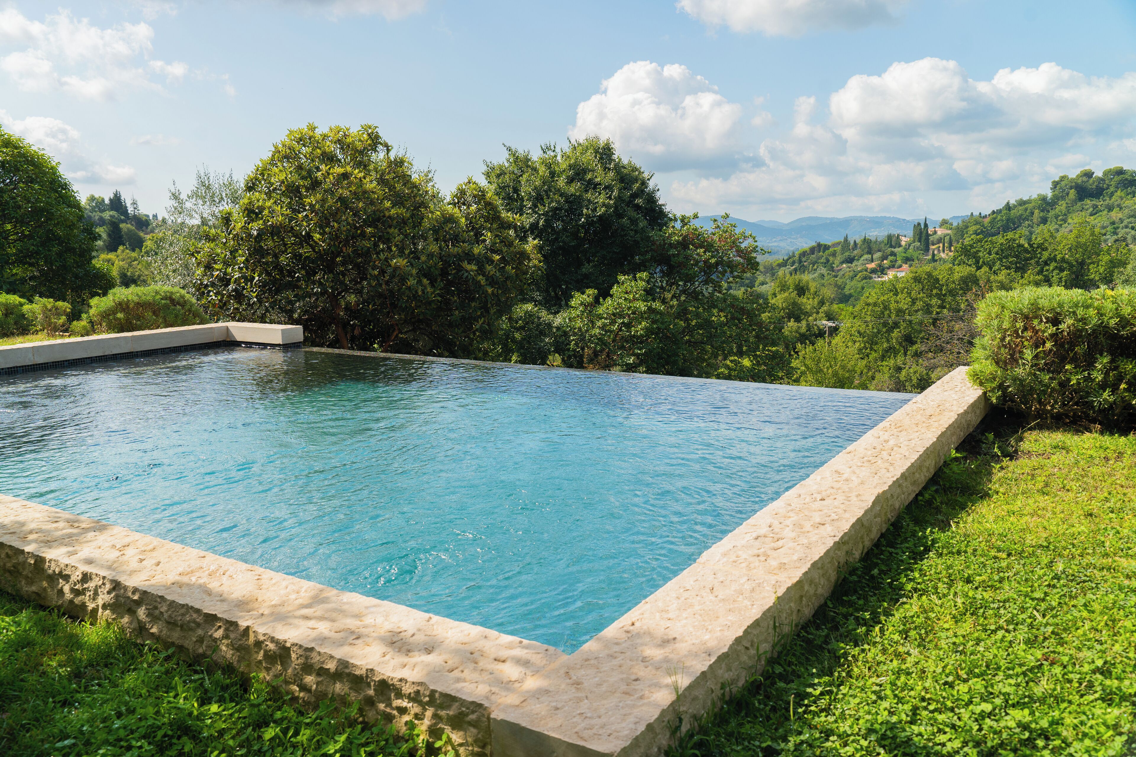Uma piscina retangular com borda de pedra, rodeada por vegetação exuberante e árvores, com vista para colinas distantes.