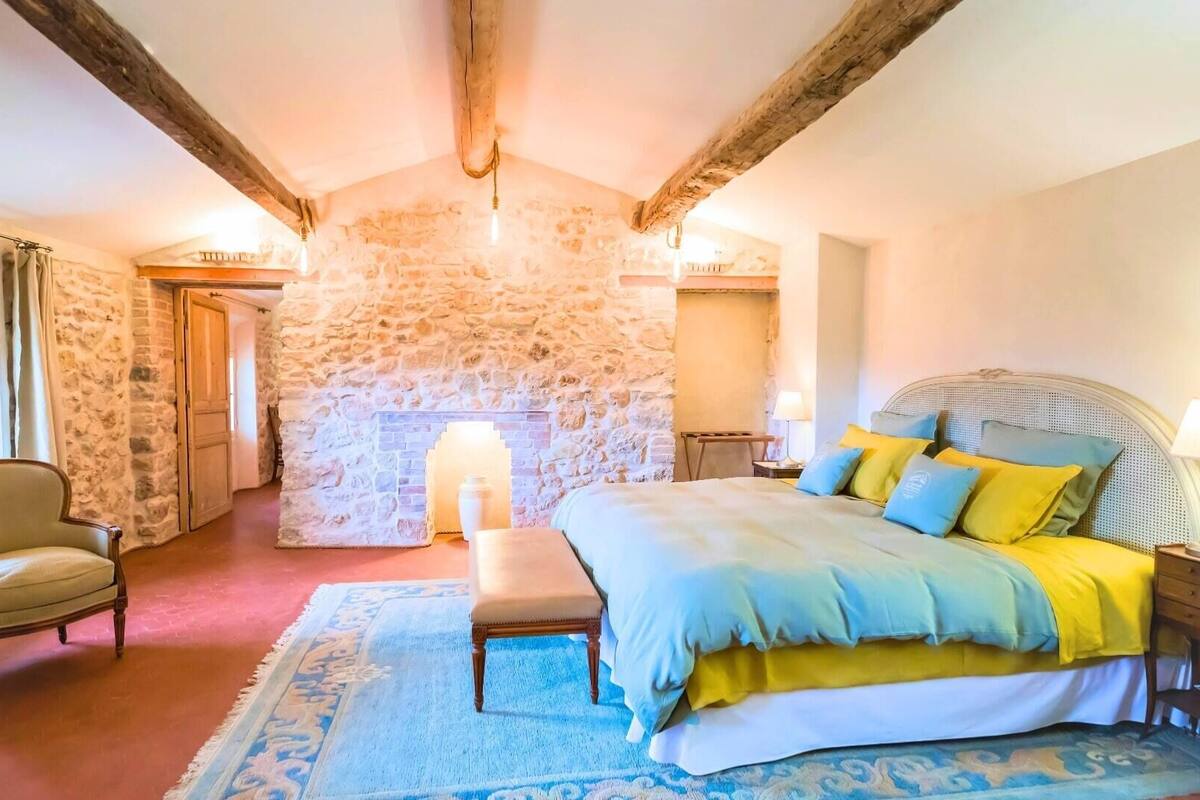 Um quarto com lareira de pedra, uma cama com várias almofadas, uma cadeira e uma mesa de cabeceira com abajur.