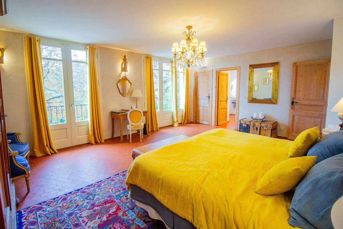 Quarto com uma cama grande, uma escrivaninha e uma cadeira. Há cortinas nas janelas, um candelabro e um espelho na parede.