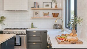 Uma cozinha moderna com forno embutido, fogão a gás, armários escuros e bancada de mármore. Há prateleiras com livros e objetos decorativos, e uma janela que traz luz natural.