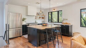 Uma cozinha moderna com geladeira de aço inoxidável, ilha de madeira com banquetas e uma grande janela com vista para vegetação.