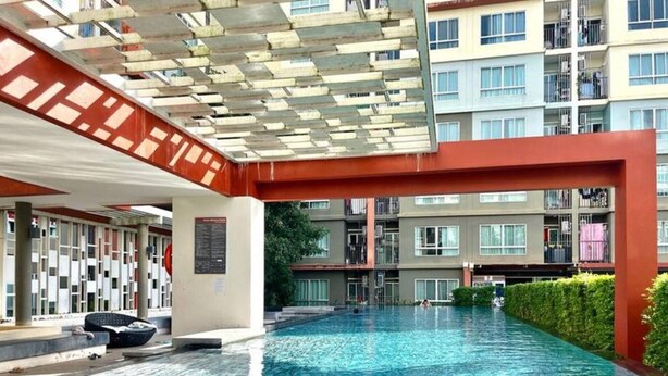 Um edifício moderno com piscina, área coberta com assentos e um prédio residencial em vários andares ao fundo.