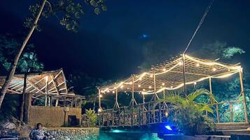 Uma área de piscina ao ar livre à noite, com pérgola de madeira e luzes de festa, cercada por paredes de pedra e plantas tropicais.