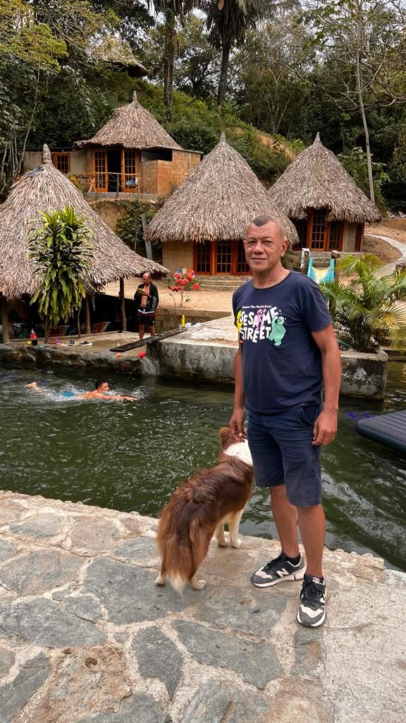 Um homem está em uma plataforma de pedra com um cachorro, uma piscina e palafitas com telhado de palha ao fundo.