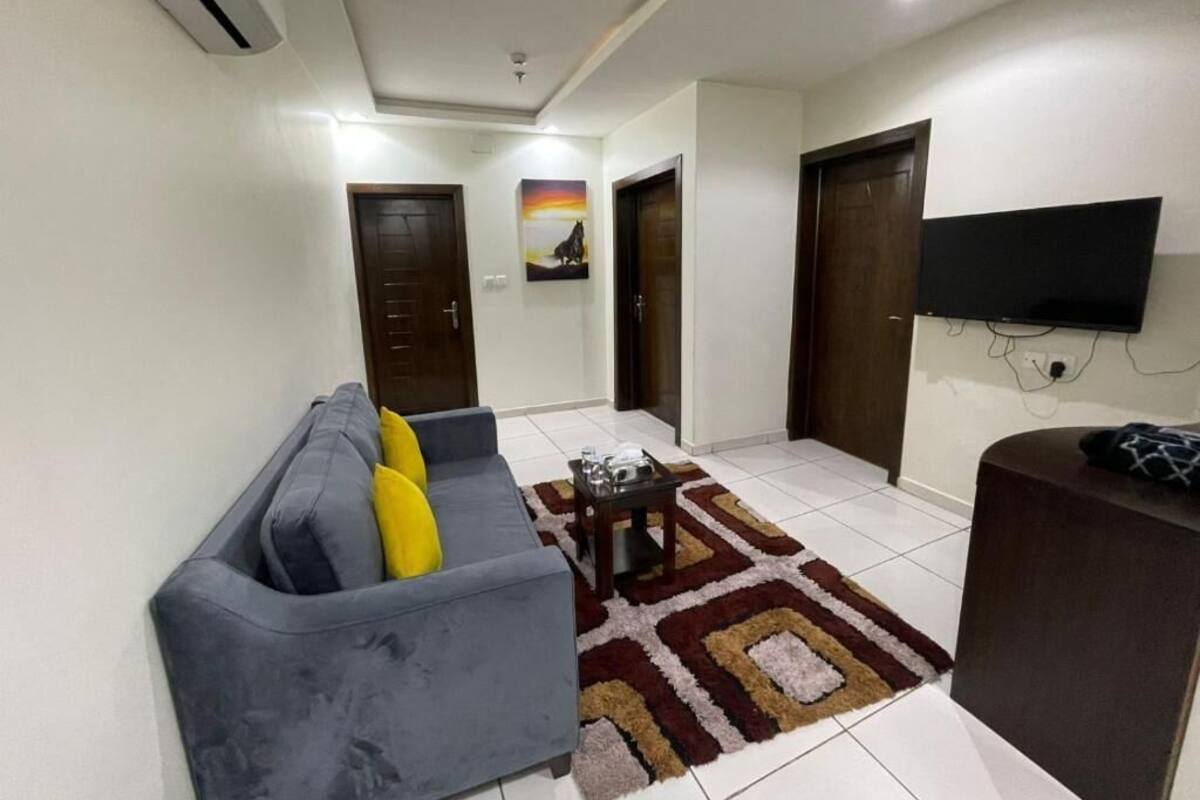 Uma sala de estar com um sofá cinza, uma mesa de centro, um tapete estampado e uma televisão fixada na parede.