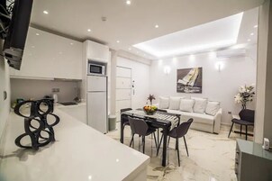 Uma cozinha moderna com armários brancos, uma área de jantar com mesa e cadeiras pretas, e um sofá branco em um espaço de estar iluminado com um claraboia.