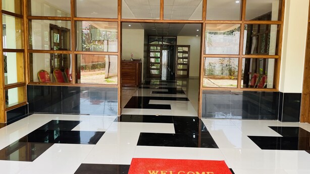 Um hall de hotel com chão em mosaico preto e branco, um tapete de boas-vindas vermelho e grandes portas de vidro com moldura de madeira que dão para a área da recepção.