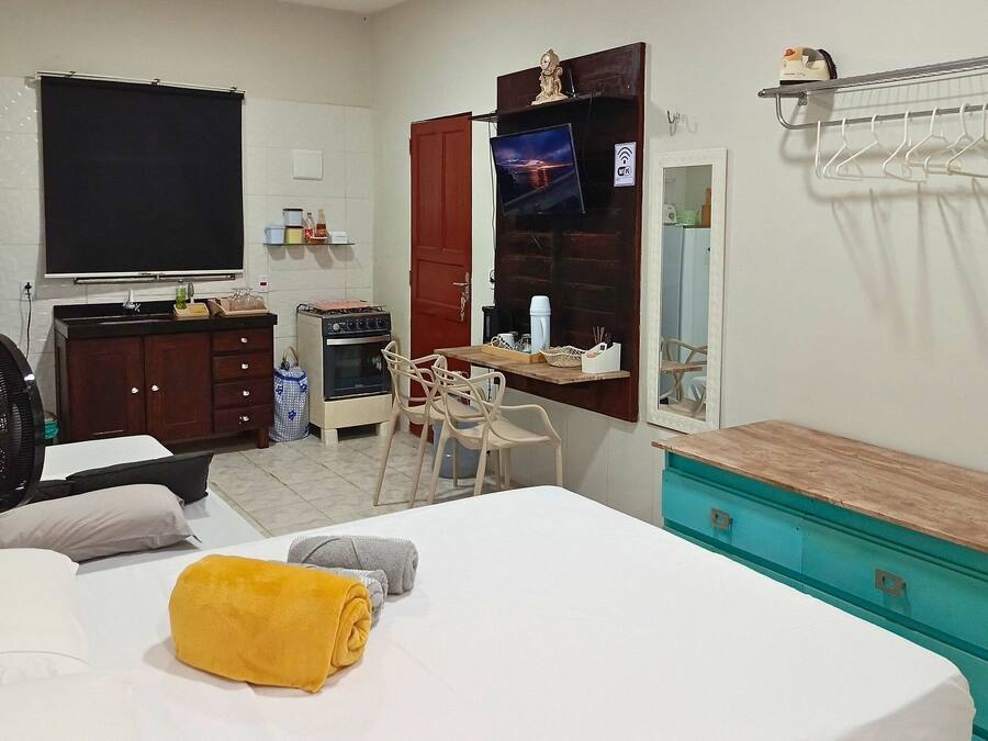 Quarto de hotel pequeno, com uma cama, escrivaninha, cadeira, televisão, guarda-roupa e uma kitchenette.