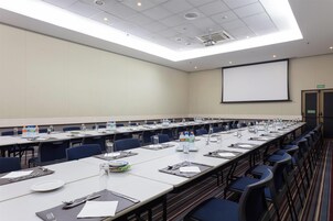 Uma sala de conferência com mesas longas, cadeiras, garrafas de água e uma tela de projetor.