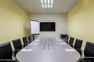 Uma sala de conferências com uma mesa comprida, cadeiras pretas, um quadro branco e uma tela de projetor.
