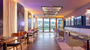 Restaurante moderno com mesas e cadeiras de madeira, uma grande porta de vidro que dá para o exterior e quadros nas paredes.