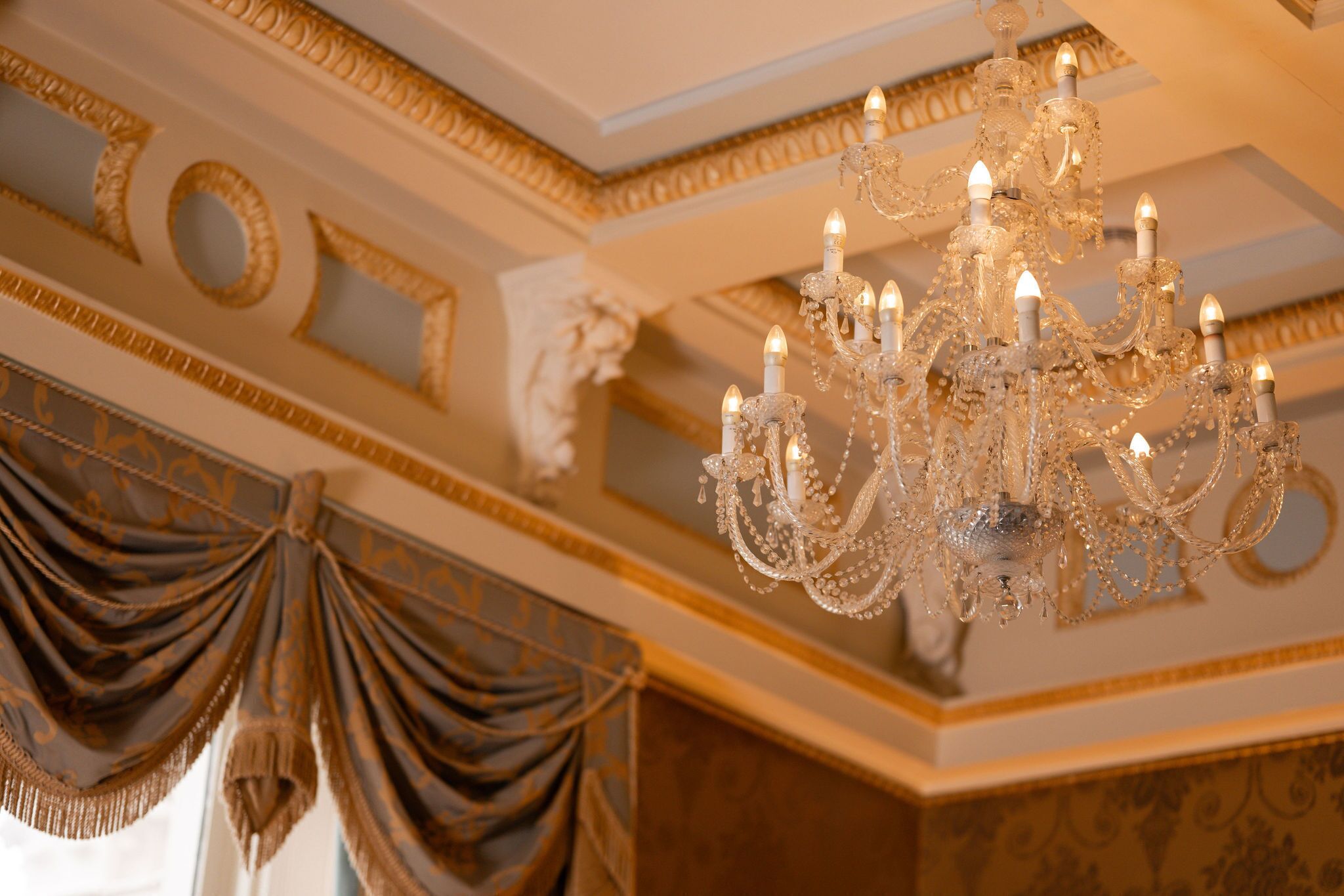 Um candelabro com várias lâmpadas que se assemelham a velas, decorações ornamentais nas paredes e cortinas com drapeados.