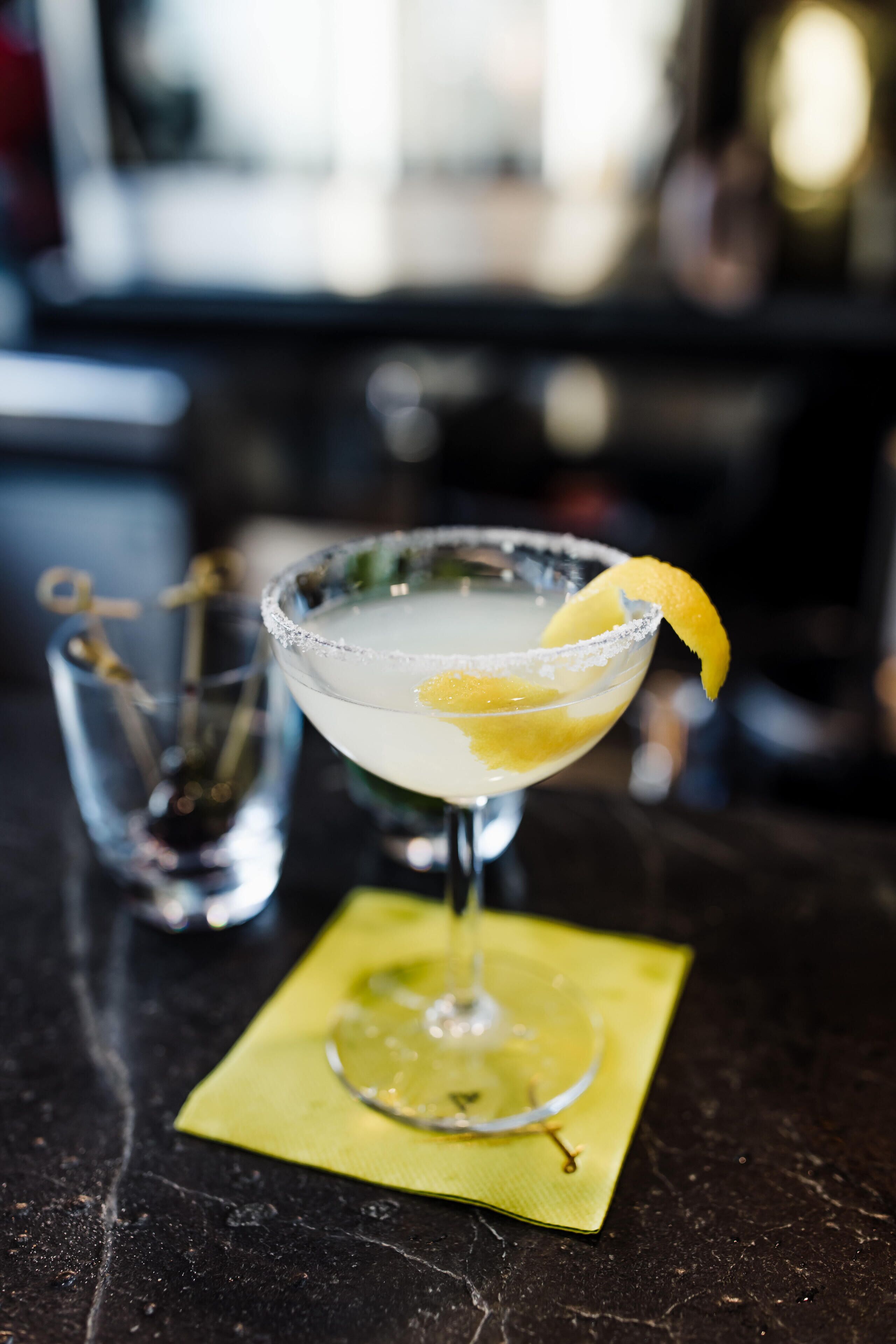 Um cocktail num copo com borda salgada e um toque de limão, apoiado sobre um guardanapo amarelo sobre uma superfície de mármore escuro.