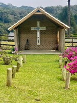 Uma pequena capela de tijolos com uma cruz, cercada por uma área gramada e postes de madeira.