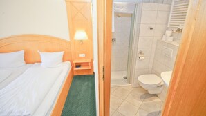 Quarto de hotel com cama, mesa de cabeceira, luminária, chuveiro e vaso sanitário.