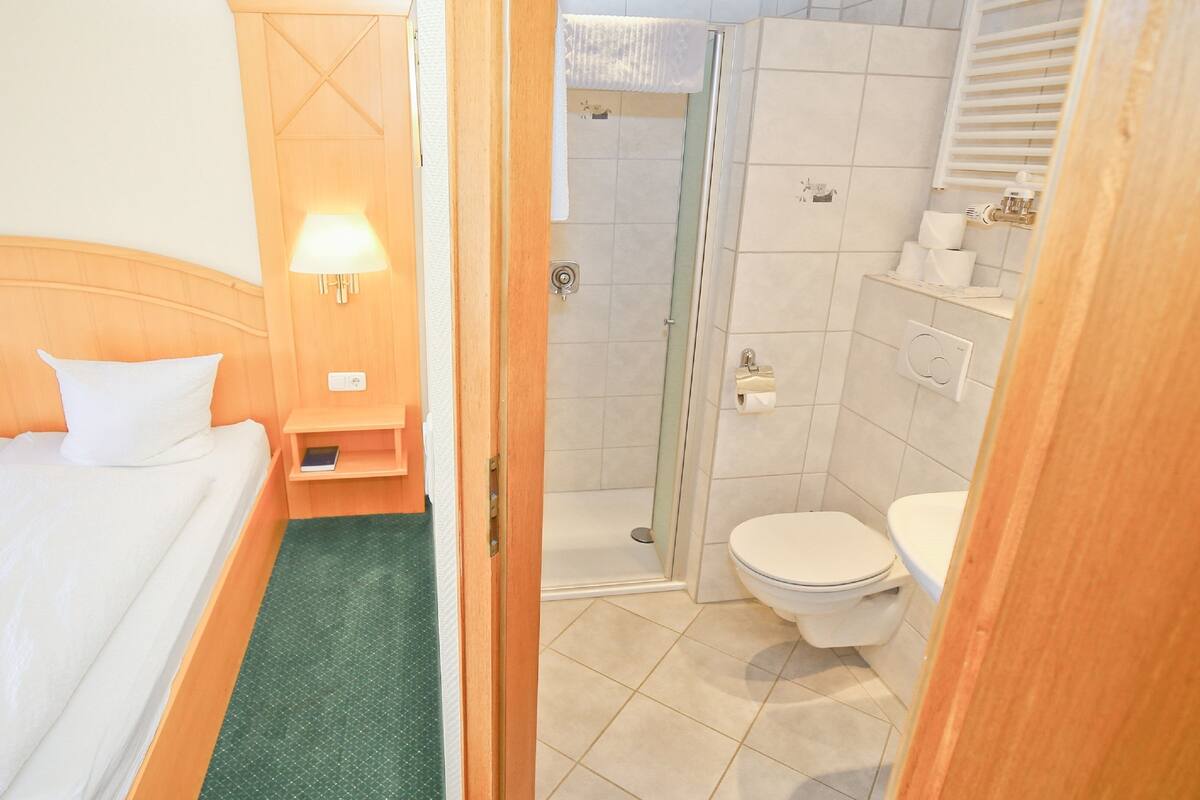 Quarto de hotel com cama, mesa de cabeceira, luminária, chuveiro e vaso sanitário.