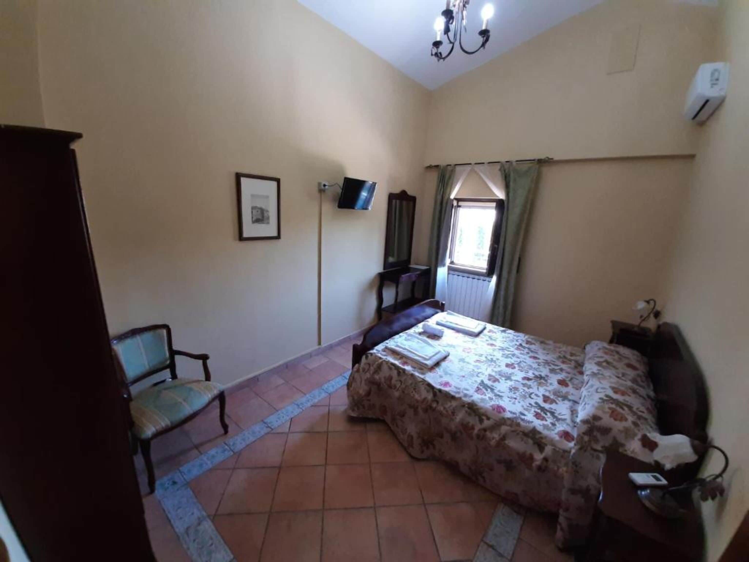 Quarto com cama, poltrona, escrivaninha, televisão e janela com cortinas.
