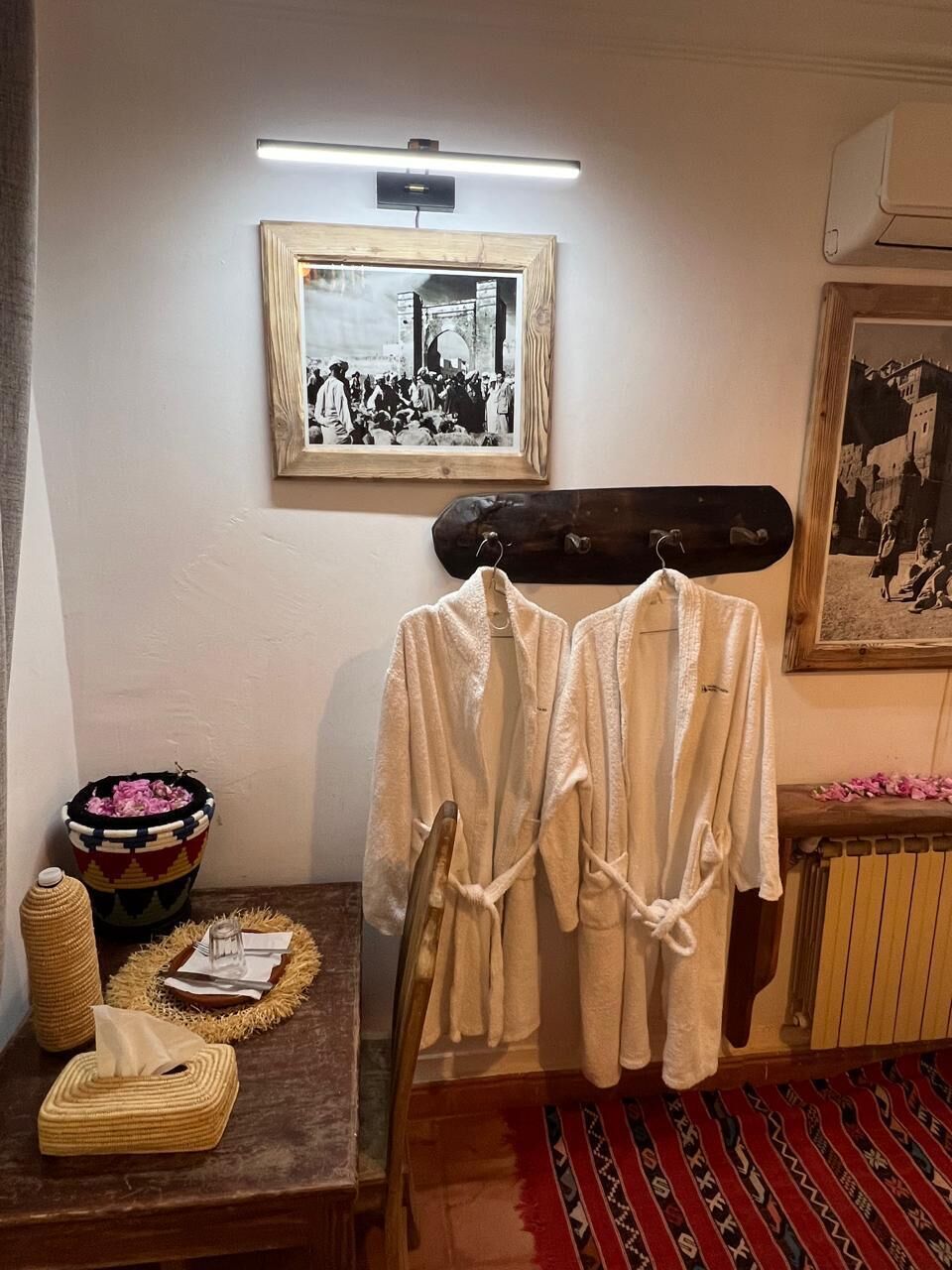 Quarto com fotografia a preto e branco emoldurada na parede, um cabideiro de madeira com roupões brancos, uma cesta de palha com flores cor-de-rosa e um tapete de palha sobre uma mesa de madeira.