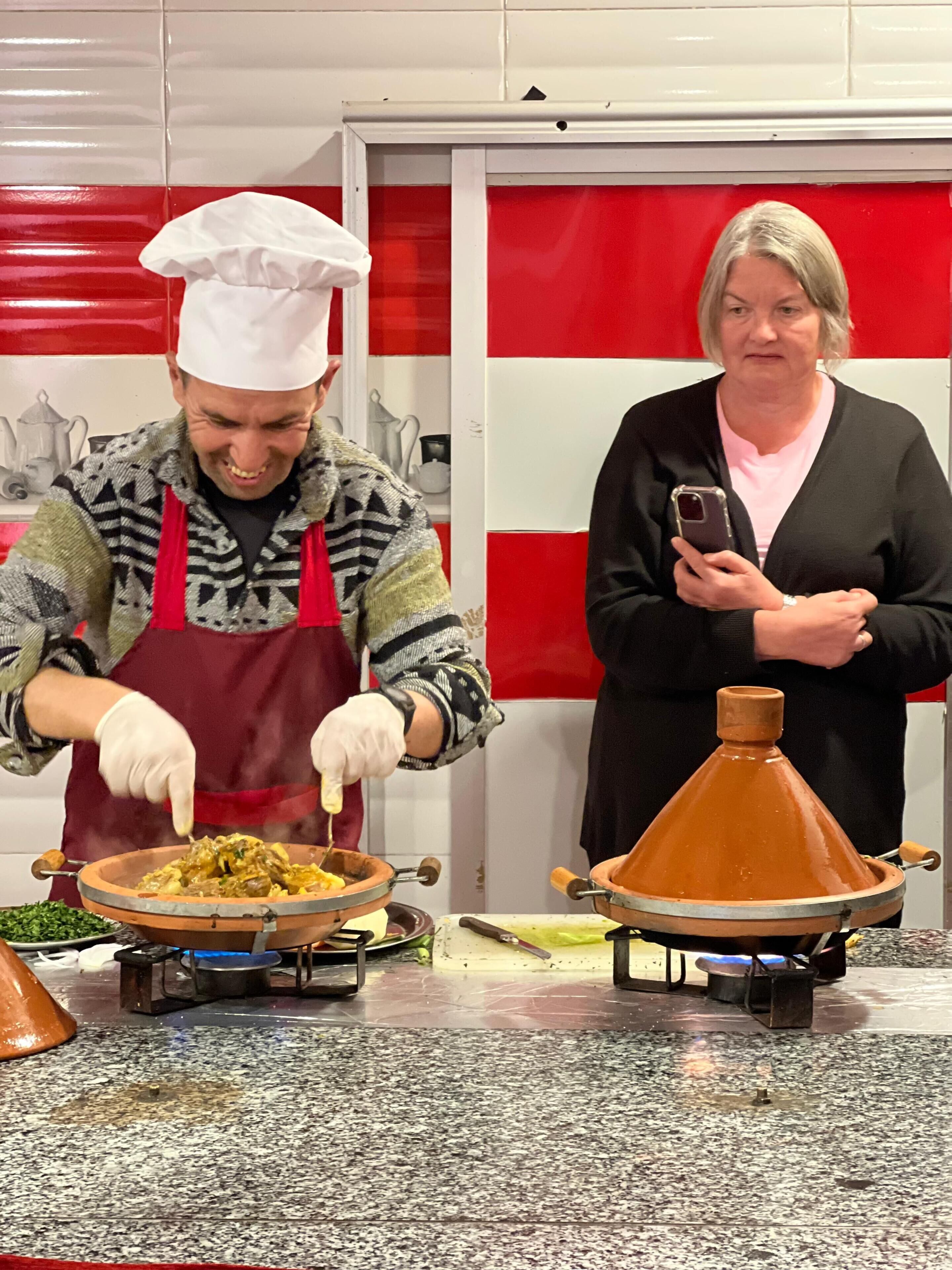 Um chef cozinha em uma cozinha com uma panela de barro grande e uma mulher observando.