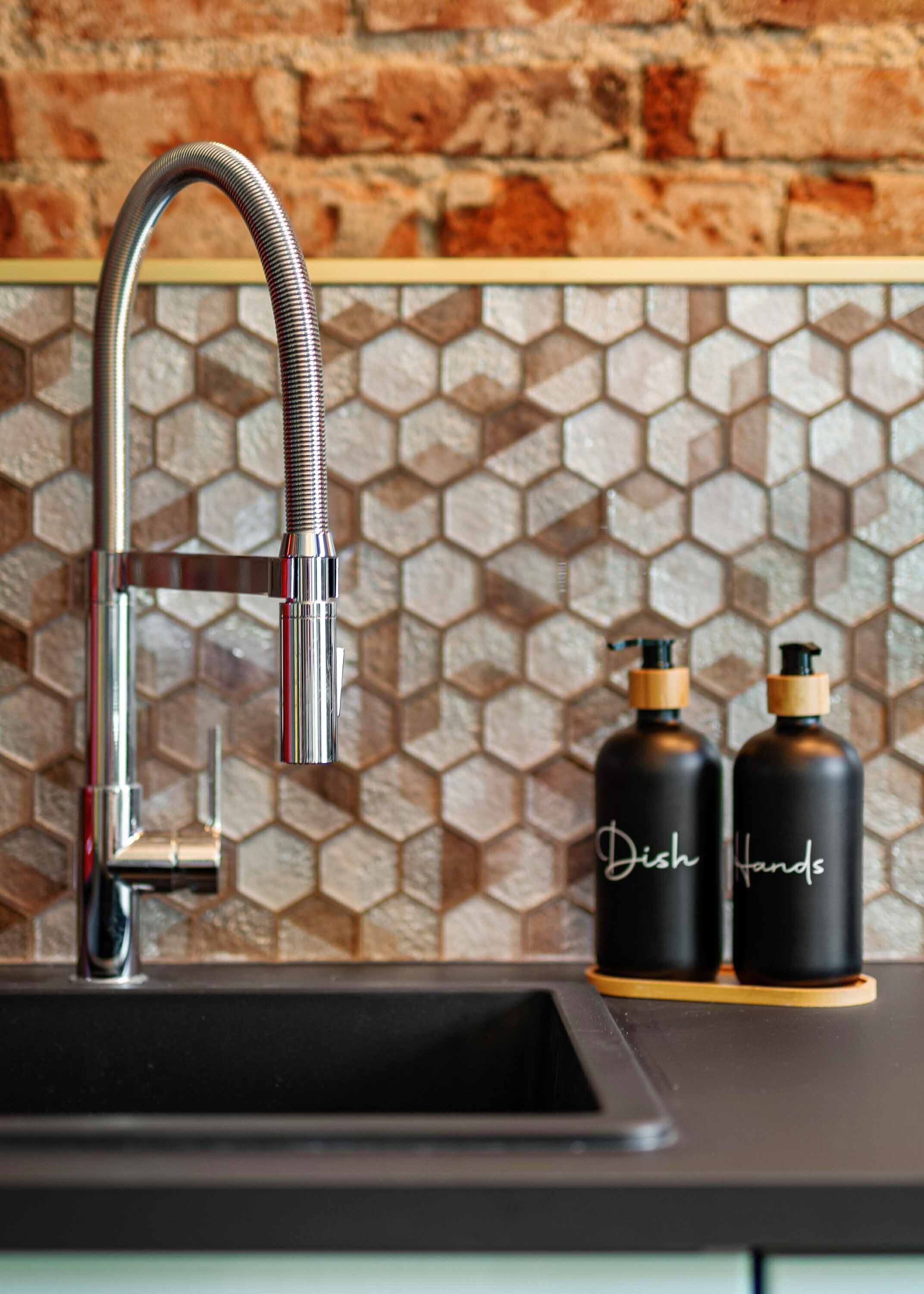 Uma pia de cozinha com uma torneira moderna, um painel de azulejos em padrão de favo de mel e duas garrafas pretas com a etiqueta "Dish Hands" em uma bancada preta.