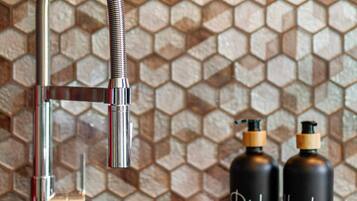 Uma pia de cozinha com uma torneira moderna, um painel de azulejos em padrão de favo de mel e duas garrafas pretas com a etiqueta "Dish Hands" em uma bancada preta.