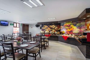 Um restaurante com várias mesas, cadeiras, um balcão de buffet e uma tela de televisão na parede.