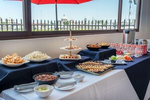 Mesa de buffet com diversos alimentos e vista para a cidade através de grandes janelas.