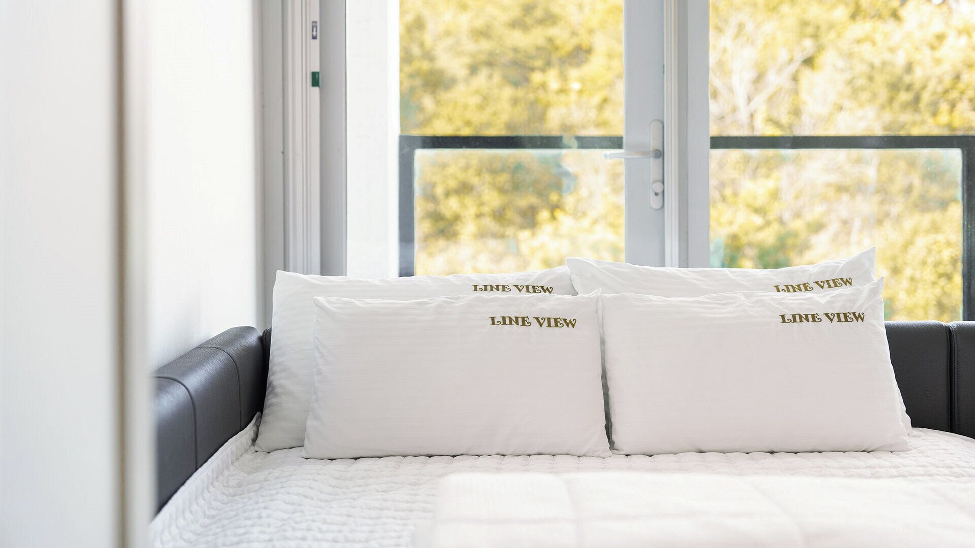 Cama com roupa de cama e almofadas brancas, janela com vista para árvores e cabeceira preta.