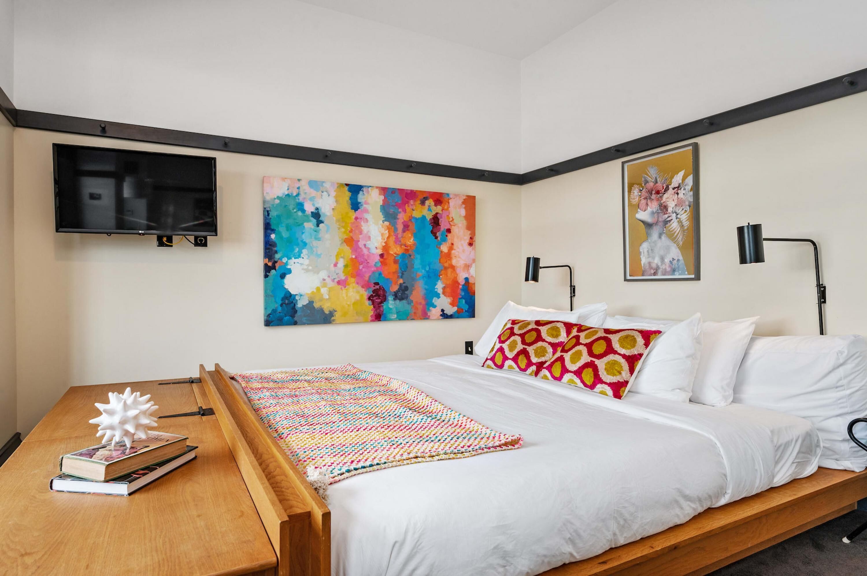 Quarto de hotel moderno com cama, televisão de tela plana, pintura abstrata colorida e obra de arte floral emoldurada.