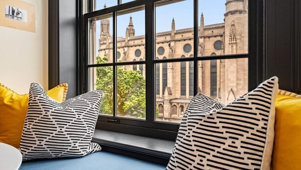 Assento junto à janela com almofadas estampadas e vista para um edifício histórico.