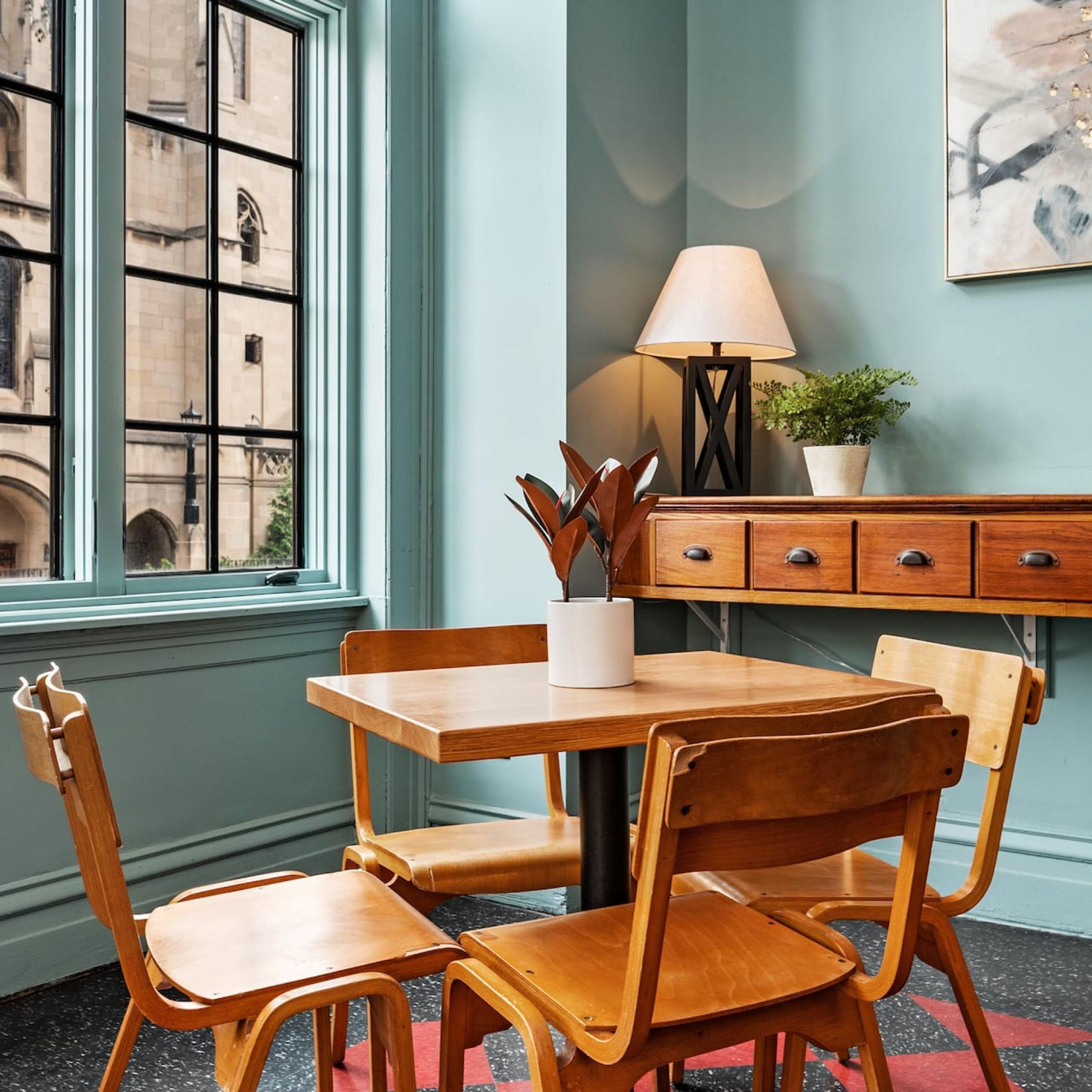 Quarto com mesa e cadeiras de madeira, abajur, planta em vaso e vista de um edifício pela janela.