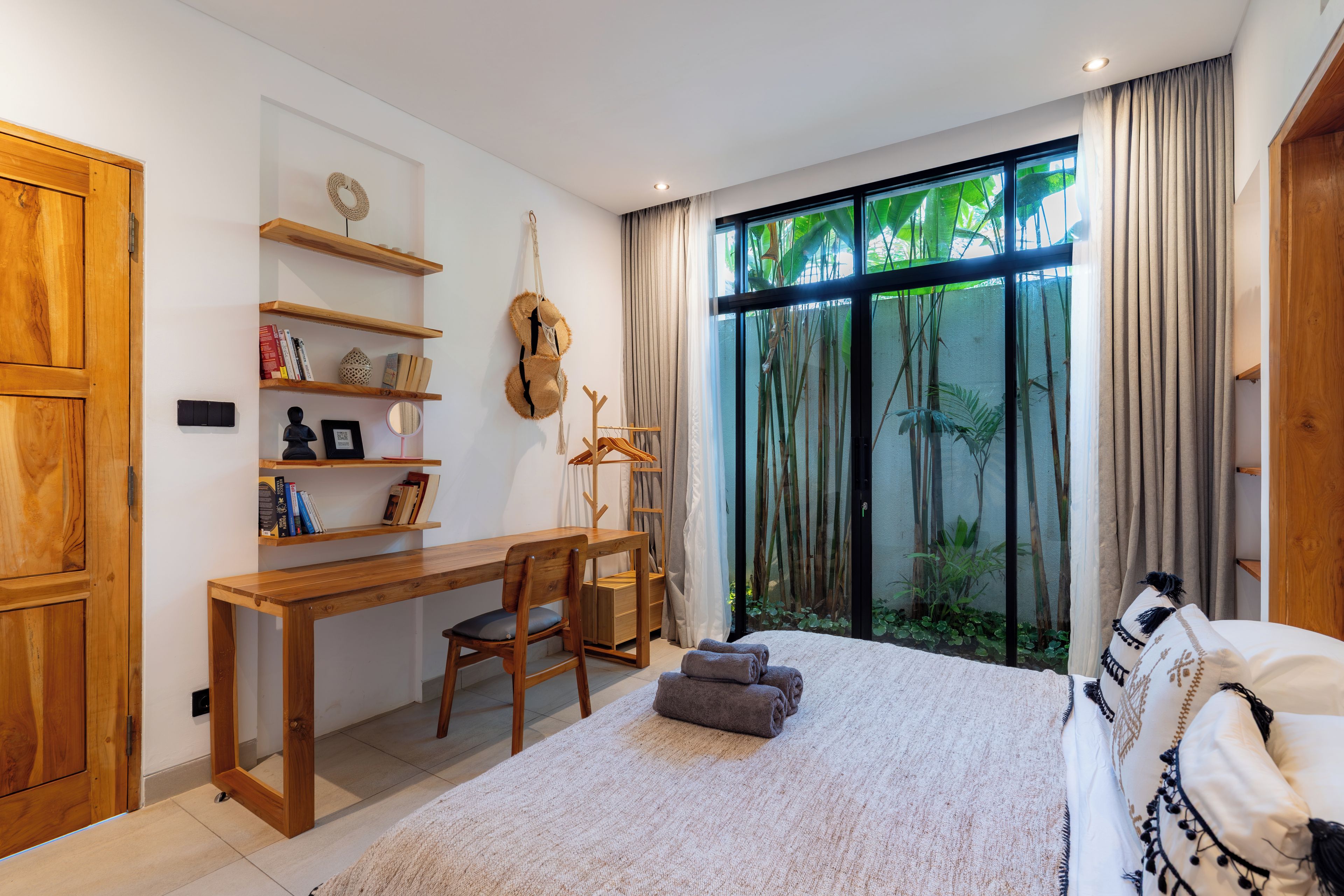 Quarto com escrivaninha de madeira, estantes de livros, cama com vista para o verde através de grandes janelas e guarda-roupa de madeira.