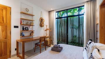 Quarto com escrivaninha de madeira, estantes de livros, cama com vista para o verde através de grandes janelas e guarda-roupa de madeira.