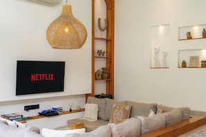 Uma sala de estar com TV de tela plana exibindo 'NETFLIX', luminária trançada, prateleira de madeira com objetos decorativos e sofá cinza.