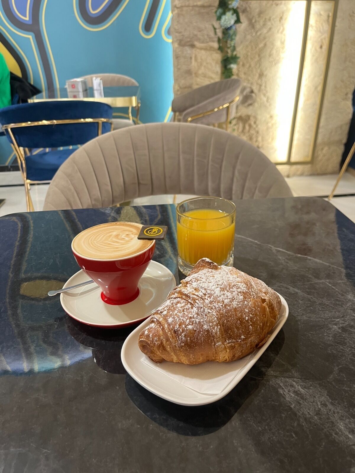 Uma mesa com um croissant e uma xícara de café, um copo de suco de laranja e uma cadeira com um design azul e dourado.