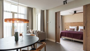 Quarto de hotel moderno com área de refeições, uma cama com cabeceira de madeira e uma grande janela com cortinas.